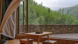 Quarto com mesa de madeira, dois bancos acolchoados, parede de pedra e vista para uma encosta arborizada.
