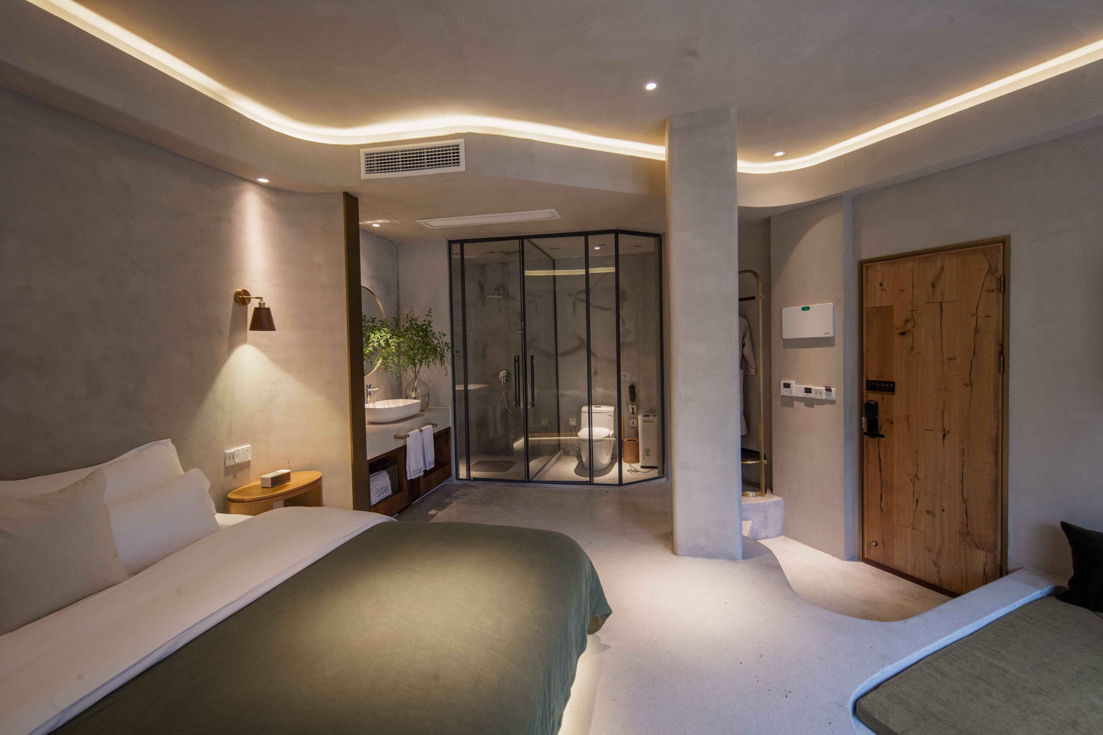 Quarto de hotel moderno com uma cama grande, área de dormir separada, banheiro com box de vidro e porta de madeira.