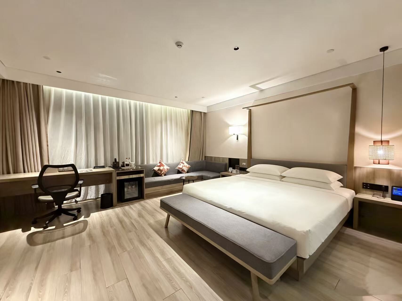Quarto de hotel moderno com uma cama grande, uma escrivaninha com cadeira, um banco, uma televisão e uma mesa de cabeceira com abajur.
