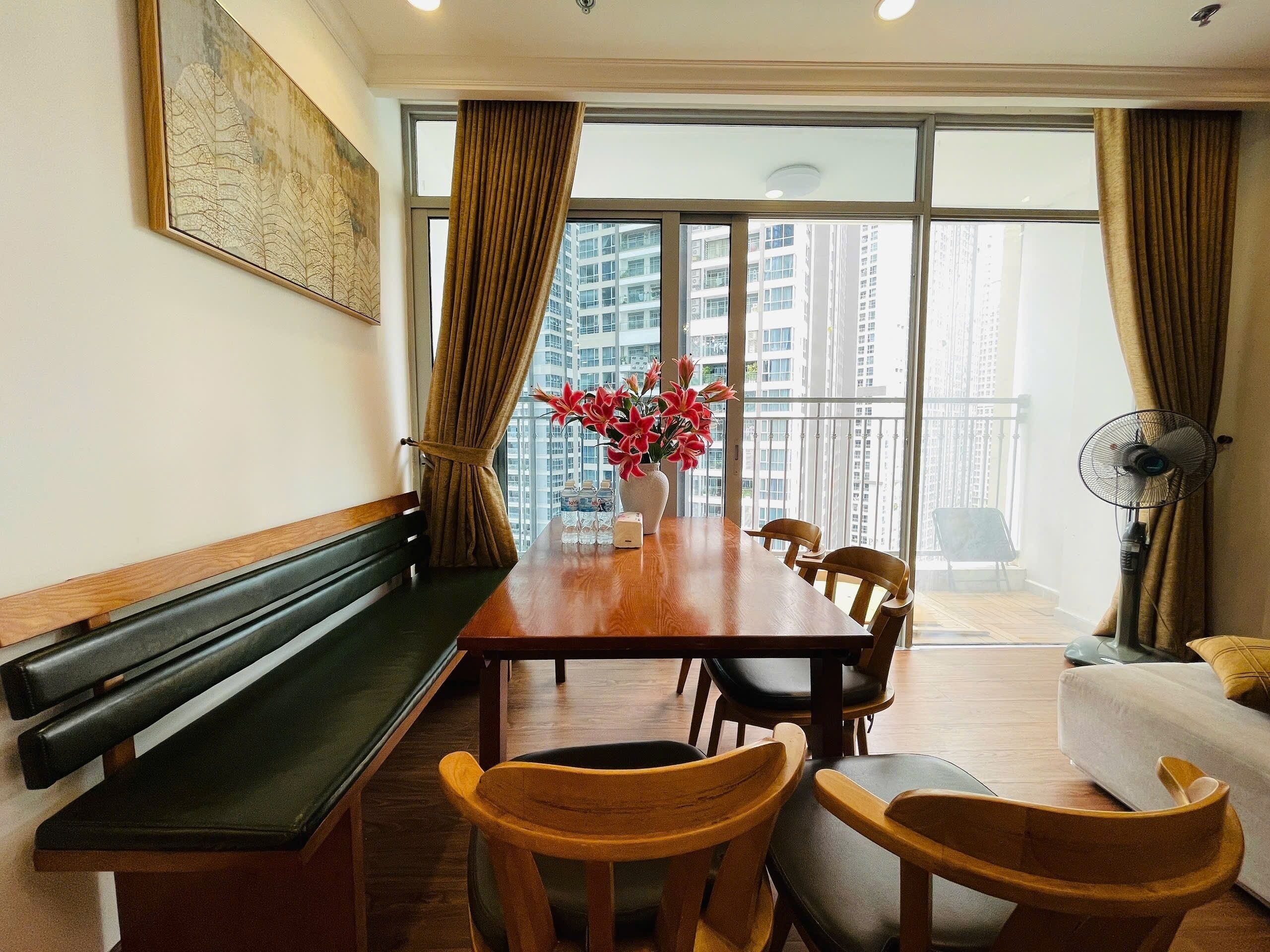 Uma área de refeições com mesa de madeira, cadeiras, um banco, um vaso com flores e uma varanda com vista para edifícios altos.