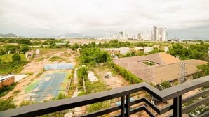Vista de uma varanda com vista para quadras esportivas, vegetação e edifícios da cidade ao longe.