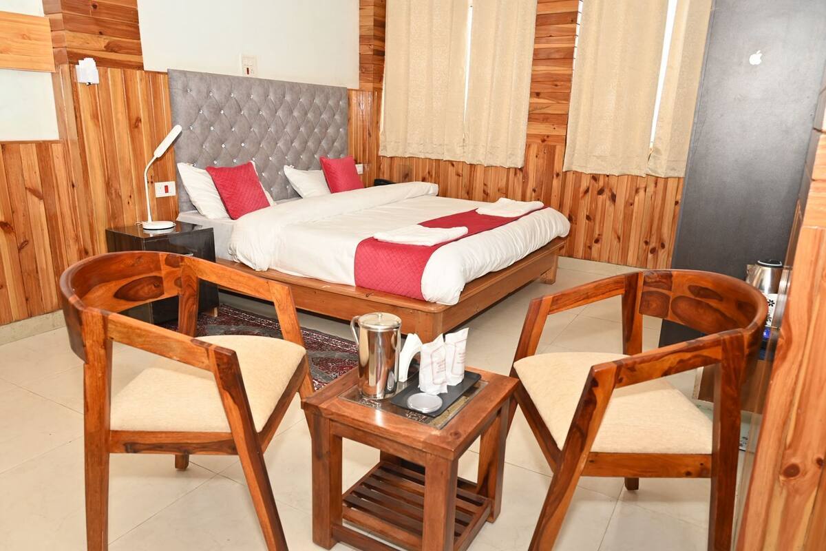 Quarto de hotel com uma cama, duas cadeiras de madeira, uma mesa pequena com um tabuleiro e uma mesinha de cabeceira com uma luminária.