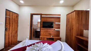 Quarto de hotel com cama, televisão fixada na parede, mobiliário de madeira e banheiro visível por uma porta aberta.