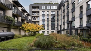 Um edifício de apartamentos moderno com varandas, uma pequena área de jardim com uma árvore e um recurso aquático visível.