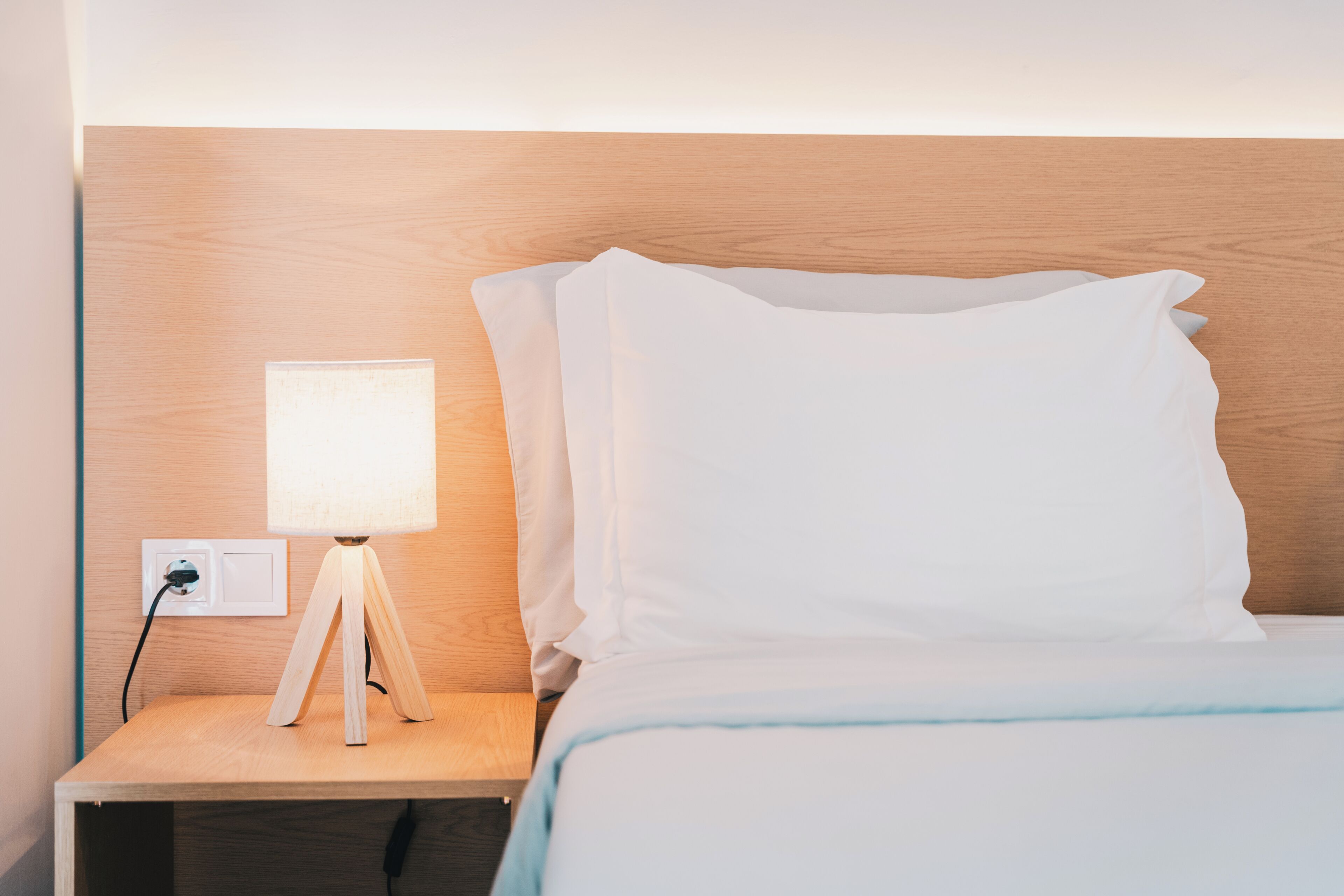Cama bem arrumada com almofadas brancas e um edredom azul, luminária de cabeceira em mesa de madeira e uma tomada na parede.