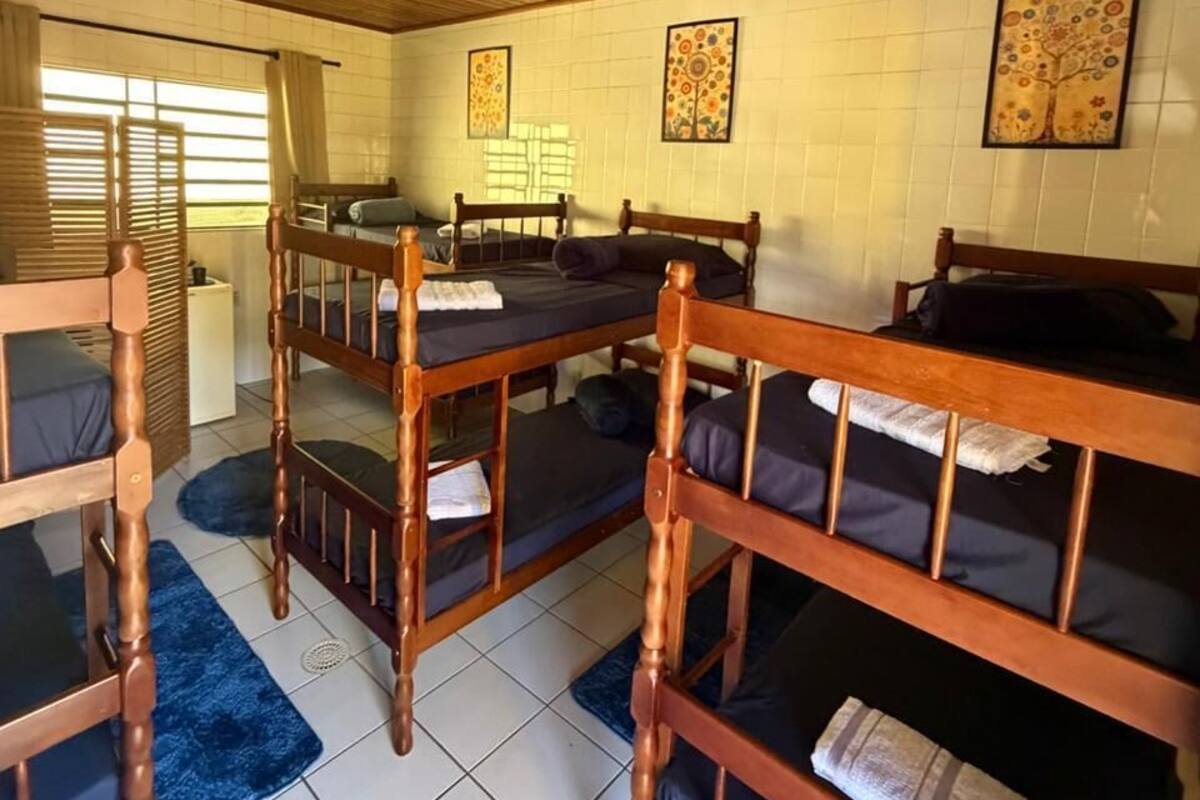 Quarto tipo dormitório com beliches, piso ladrilhado, teto de madeira e uma janela com persianas.