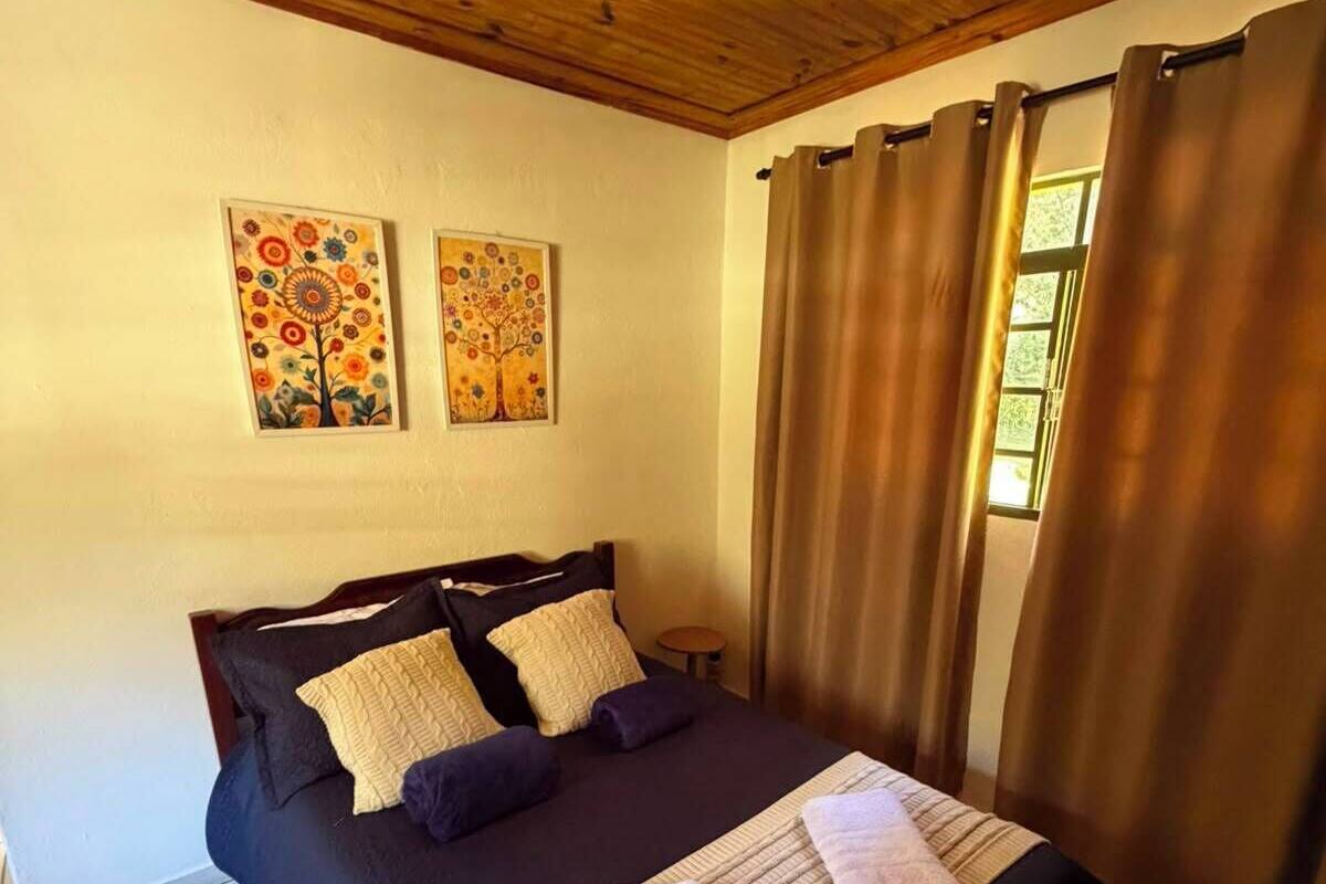 Quarto com teto de madeira, cama com cobertura escura, duas obras de arte nas paredes e janela com cortinas.