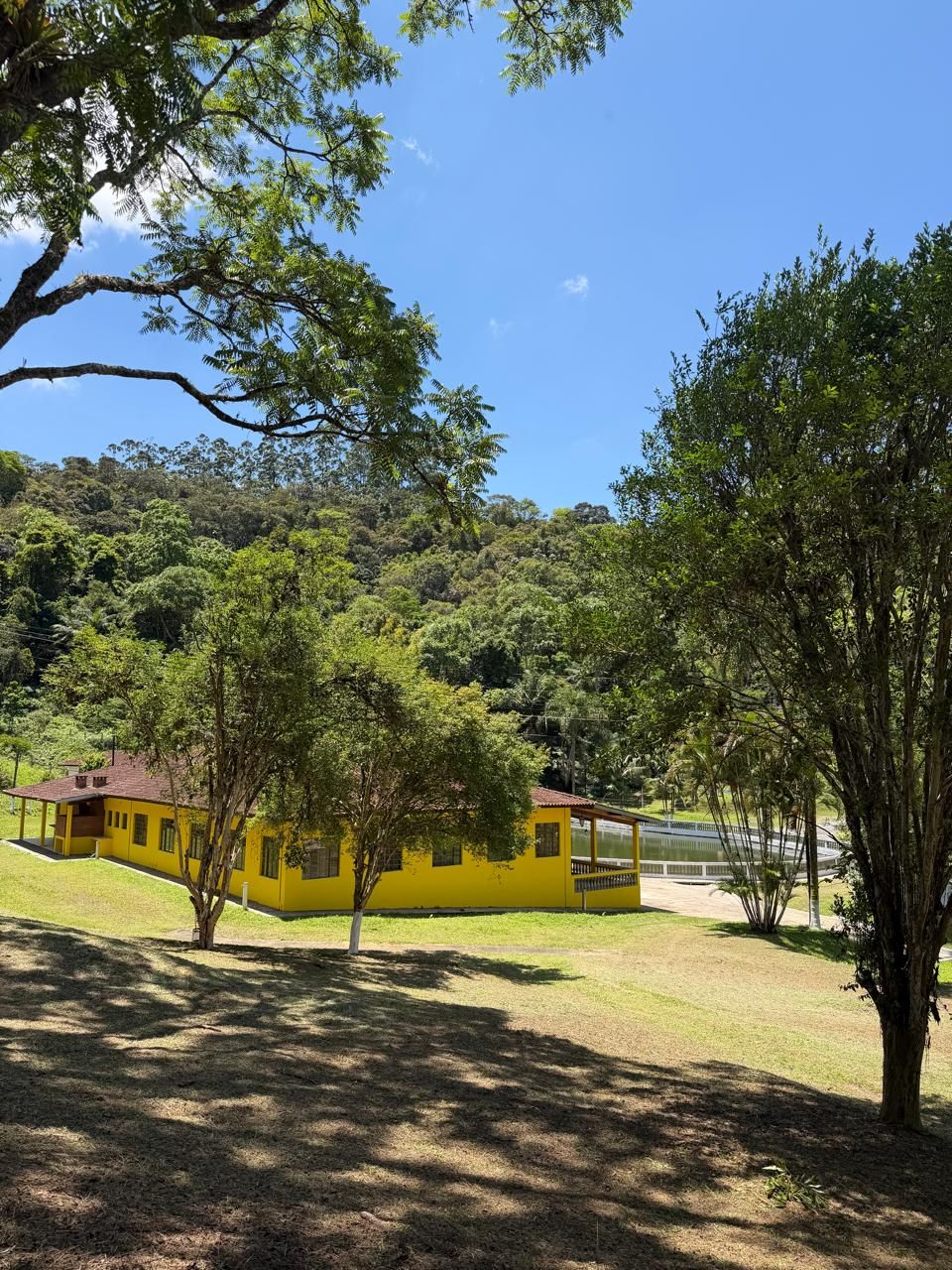 Um edifício amarelo com telhado vermelho, rodeado por árvores e uma paisagem acidentada.
