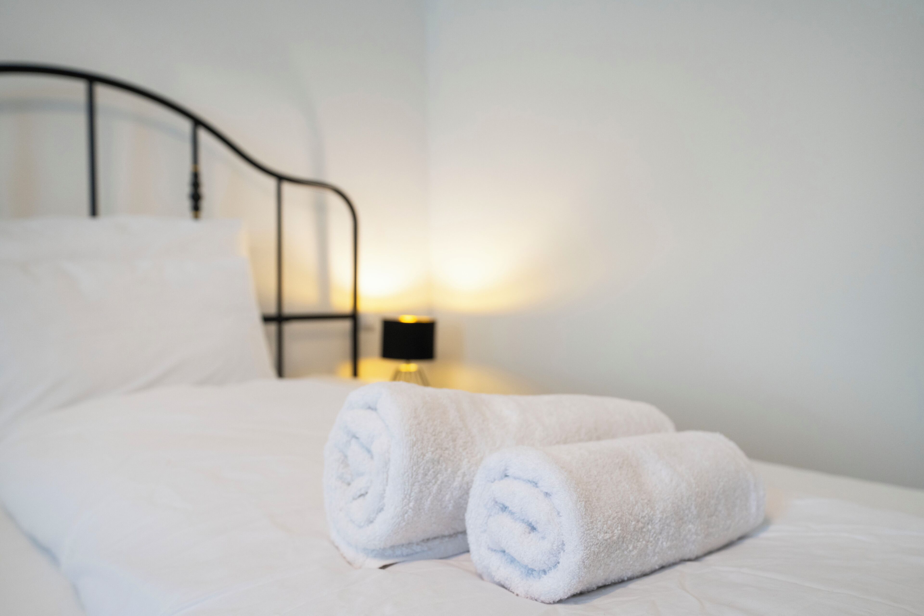 Cama bem arrumada com duas toalhas brancas enroladas e cabeceira de metal preto.