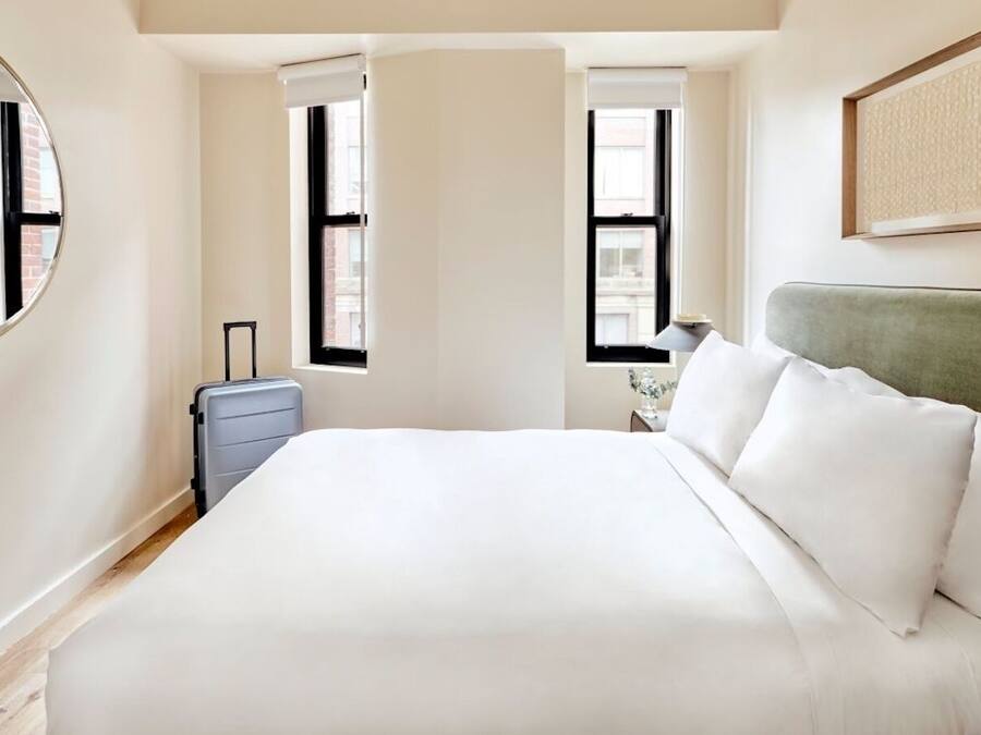 Quarto de hotel com uma cama grande, uma mala e duas janelas.