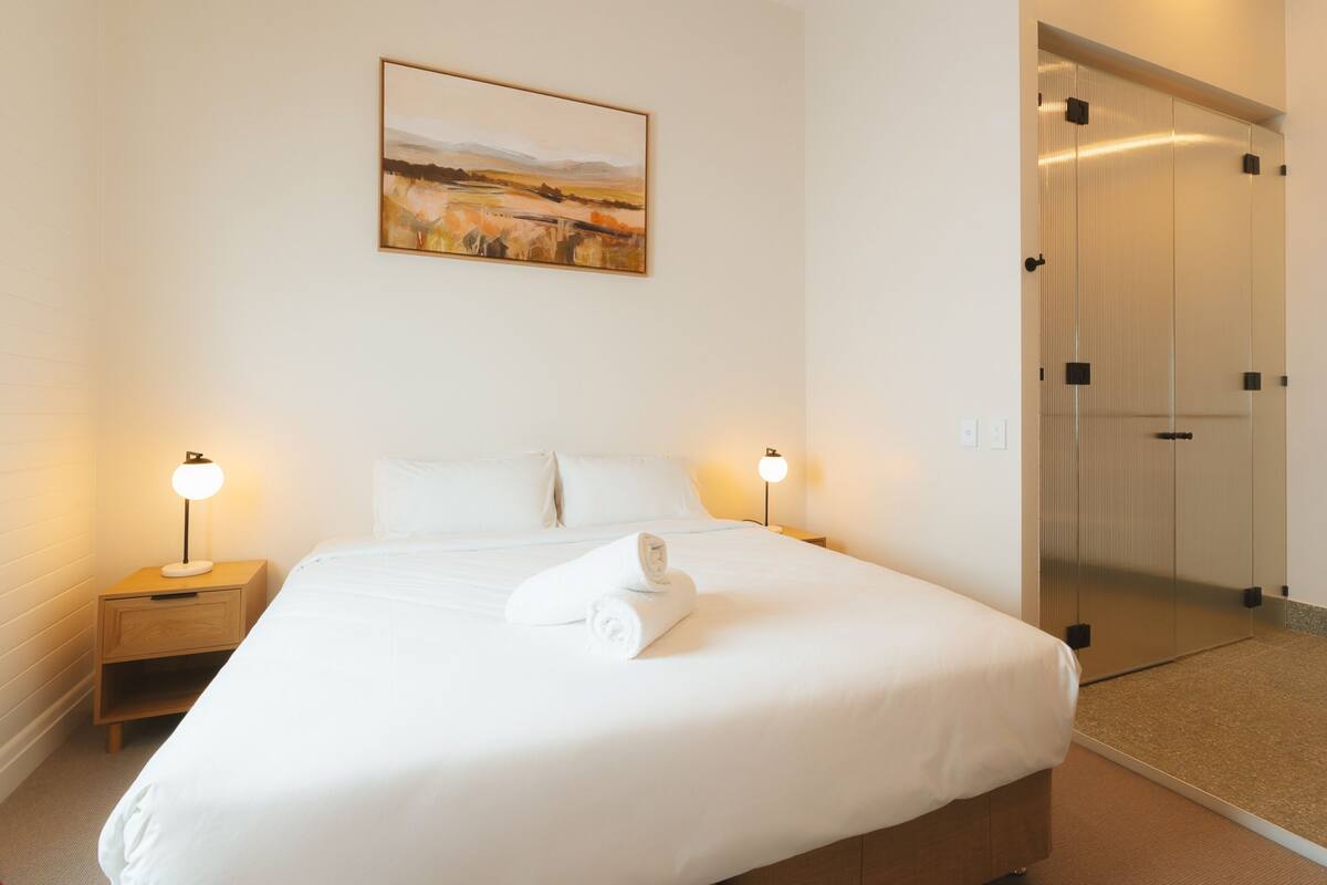 Cama bem arrumada com lençóis brancos e toalha enrolada, luminária de cabeceira, quadro na parede e porta deslizante à direita.