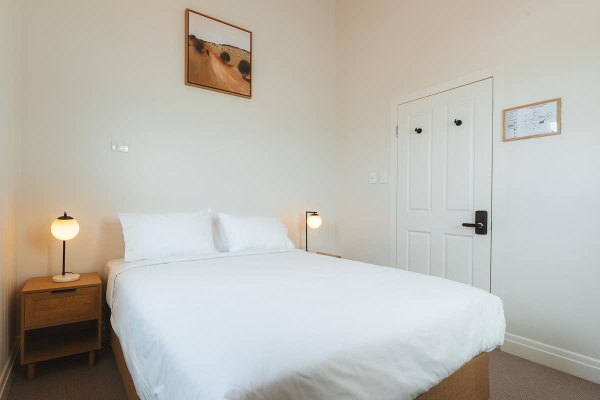 Cama bem arrumada com roupa de cama branca, mesa de cabeceira com abajur, porta com puxador e quadro na parede.