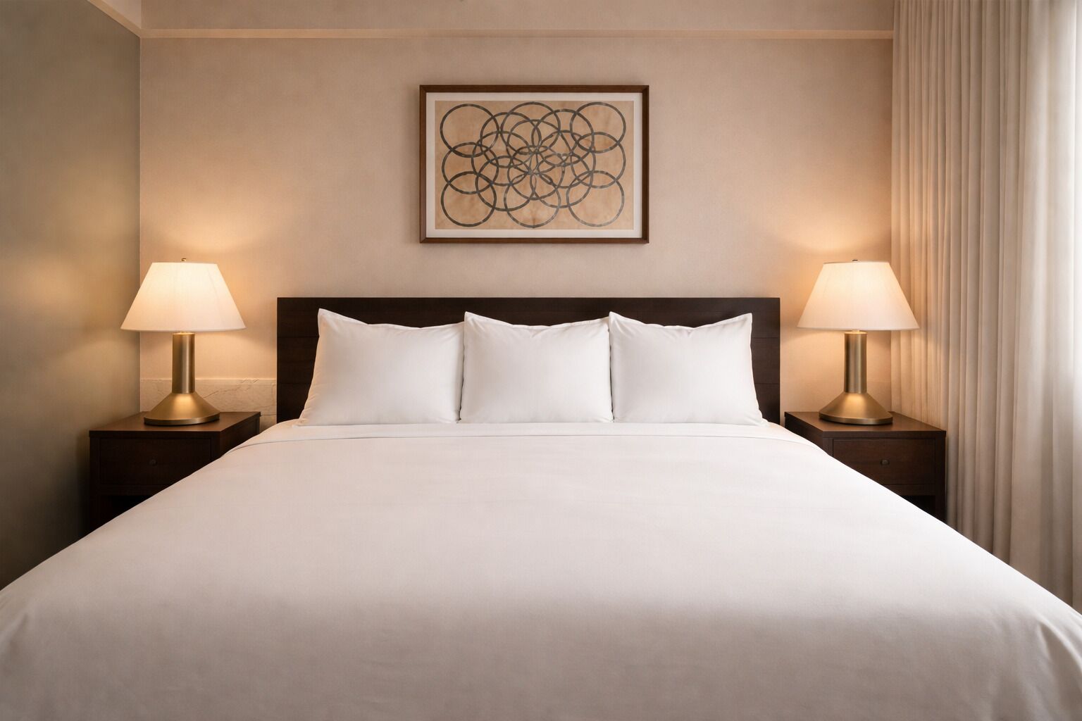 Cama bem arrumada com lençóis brancos e cabeceira escura, ladeada por duas mesinhas de cabeceira com abajures. Uma obra de arte abstrata emoldurada está centralizada acima da cama.
