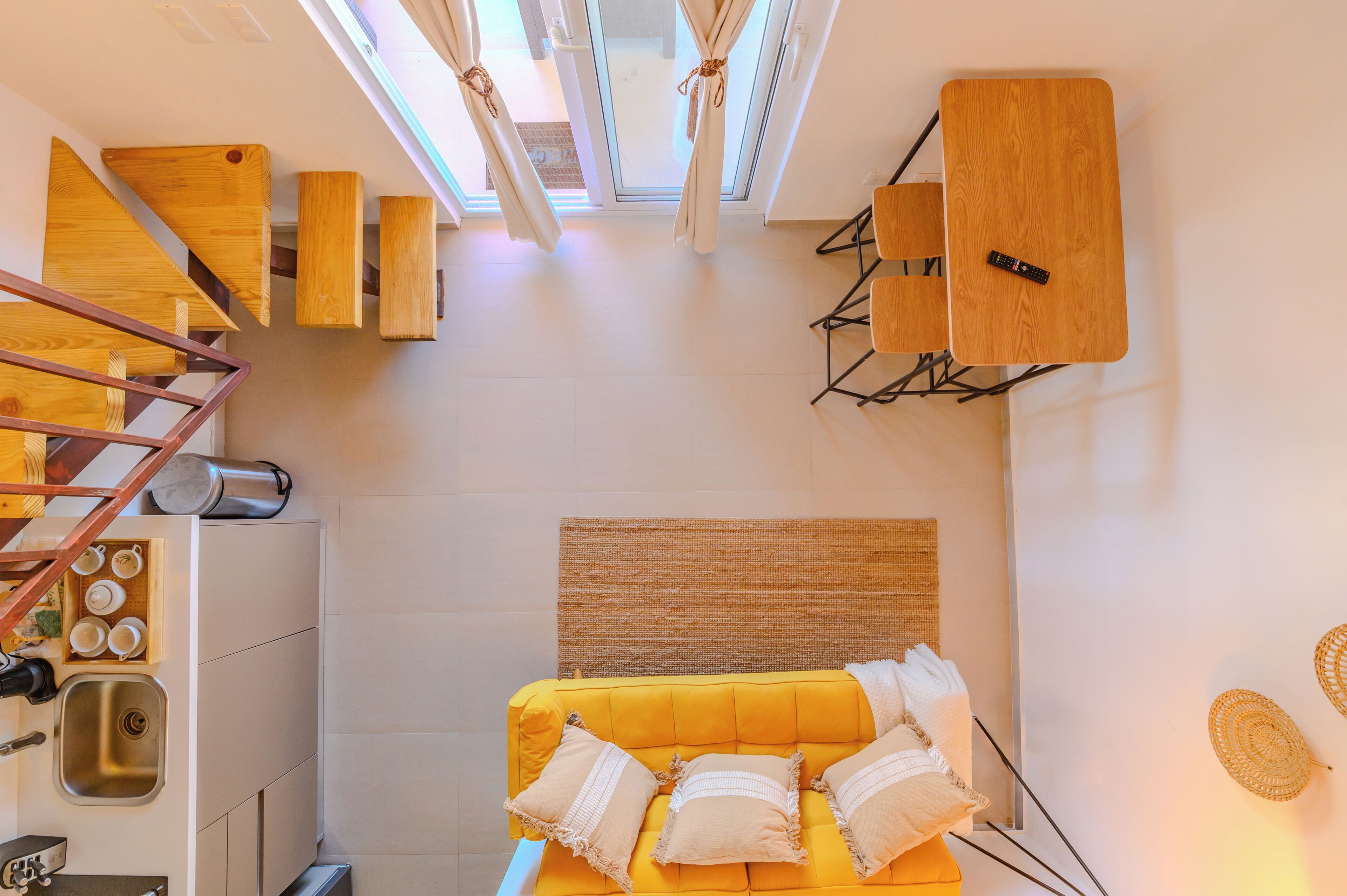 Um espaço compacto com um sofá amarelo, área de cozinha com pia e armários, escada de madeira e uma mesa pequena com um controle remoto.
