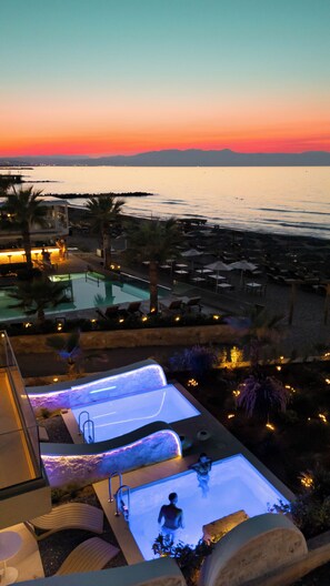 Um resort com área de piscina, espreguiçadeiras e vista para o oceano ao pôr do sol.