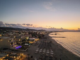 Uma vila costeira ao pôr do sol, com uma praia, fileiras de espreguiçadeiras e um porto de iates.