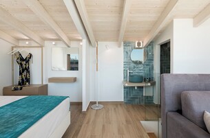 Quarto moderno com cama, cabide, espelho, banheiro com pia e espelho redondo, e sofá cinza.