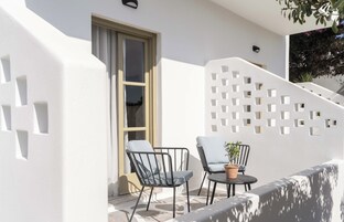 Paredes externas brancas com design geométrico recortado, uma janela e uma pequena área de estar ao ar livre com duas cadeiras e uma mesa.
