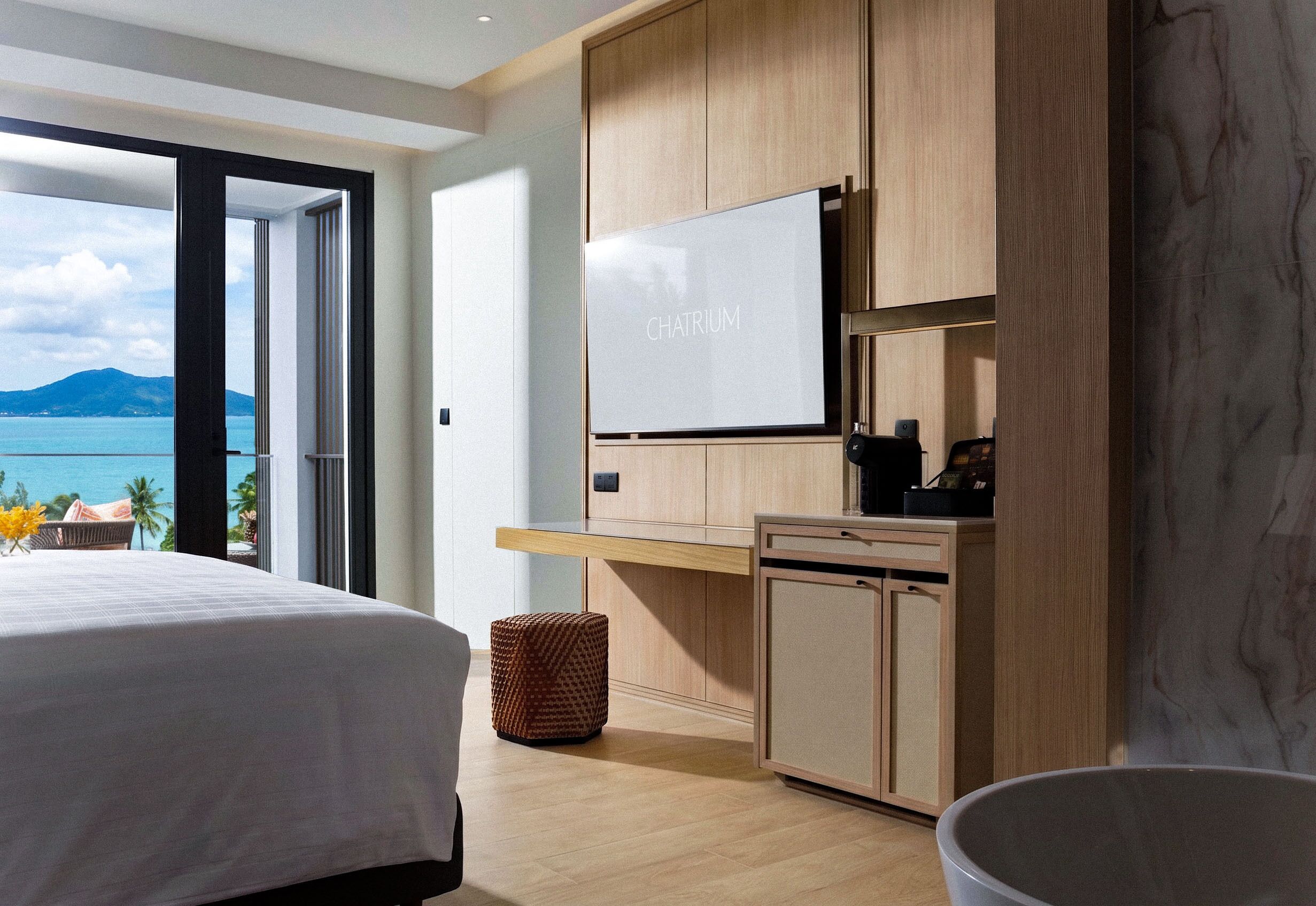 Quarto de hotel moderno com cama, cozinha compacta, uma TV grande e vista para o oceano.