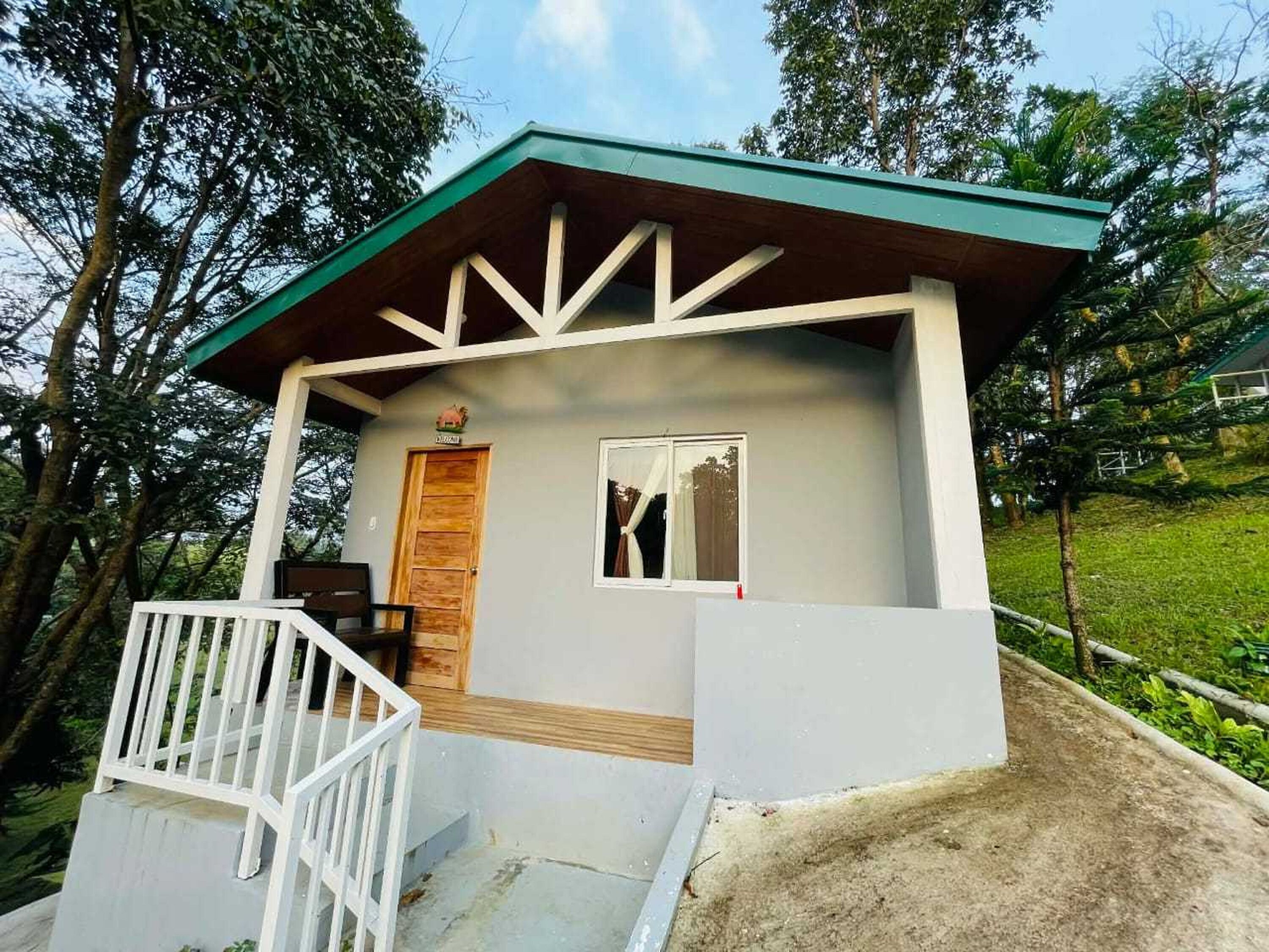 Um pequeno edifício térreo com telhado verde e paredes brancas, apresentando uma porta de madeira e uma janela.
