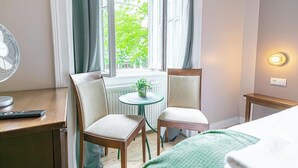 Quarto de hotel com cama, uma pequena mesa com cadeiras, um ventilador e uma janela com cortinas.