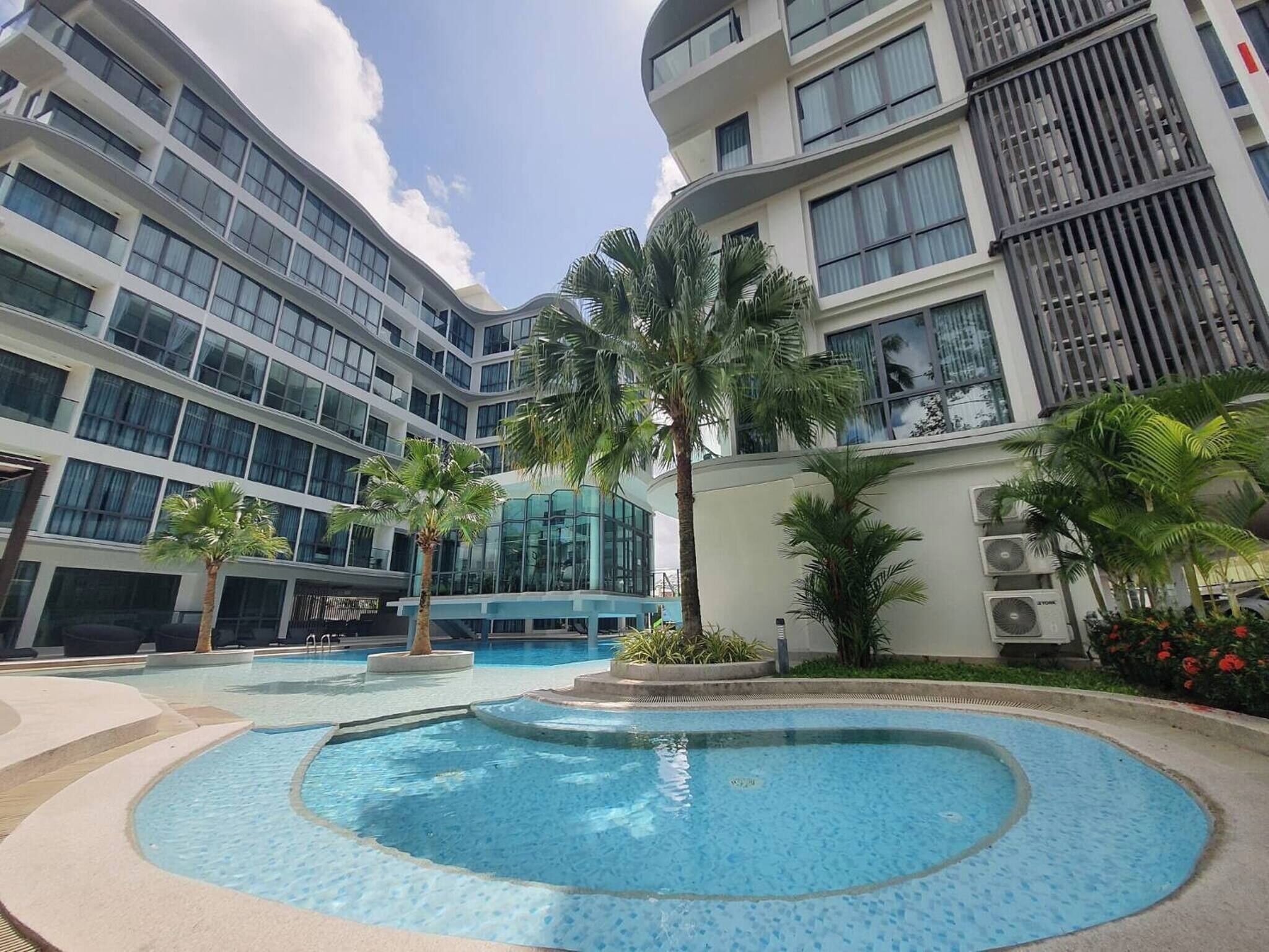 Um hotel moderno com piscina central, rodeado por palmeiras e um edifício com vários andares e varandas.