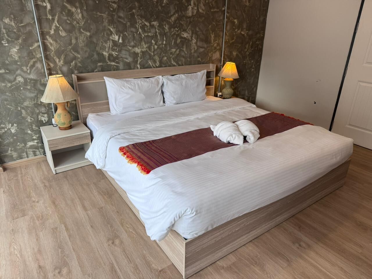Cama bem arrumada com roupa de cama branca e manta vermelha, duas luminárias de cabeceira e cabeceira de madeira.