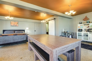 Quarto com cama, mesa de madeira, um banco, cozinha compacta e porta para outro espaço.