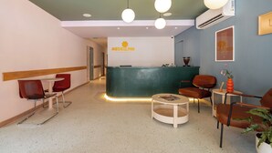 Uma área de recepção com balcão verde, cadeiras vermelhas e castanhas, uma mesa redonda e iluminação pendente.