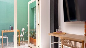 Quarto com mesa de madeira, cadeiras, televisão e porta que dá para uma varanda com plantas em vasos.