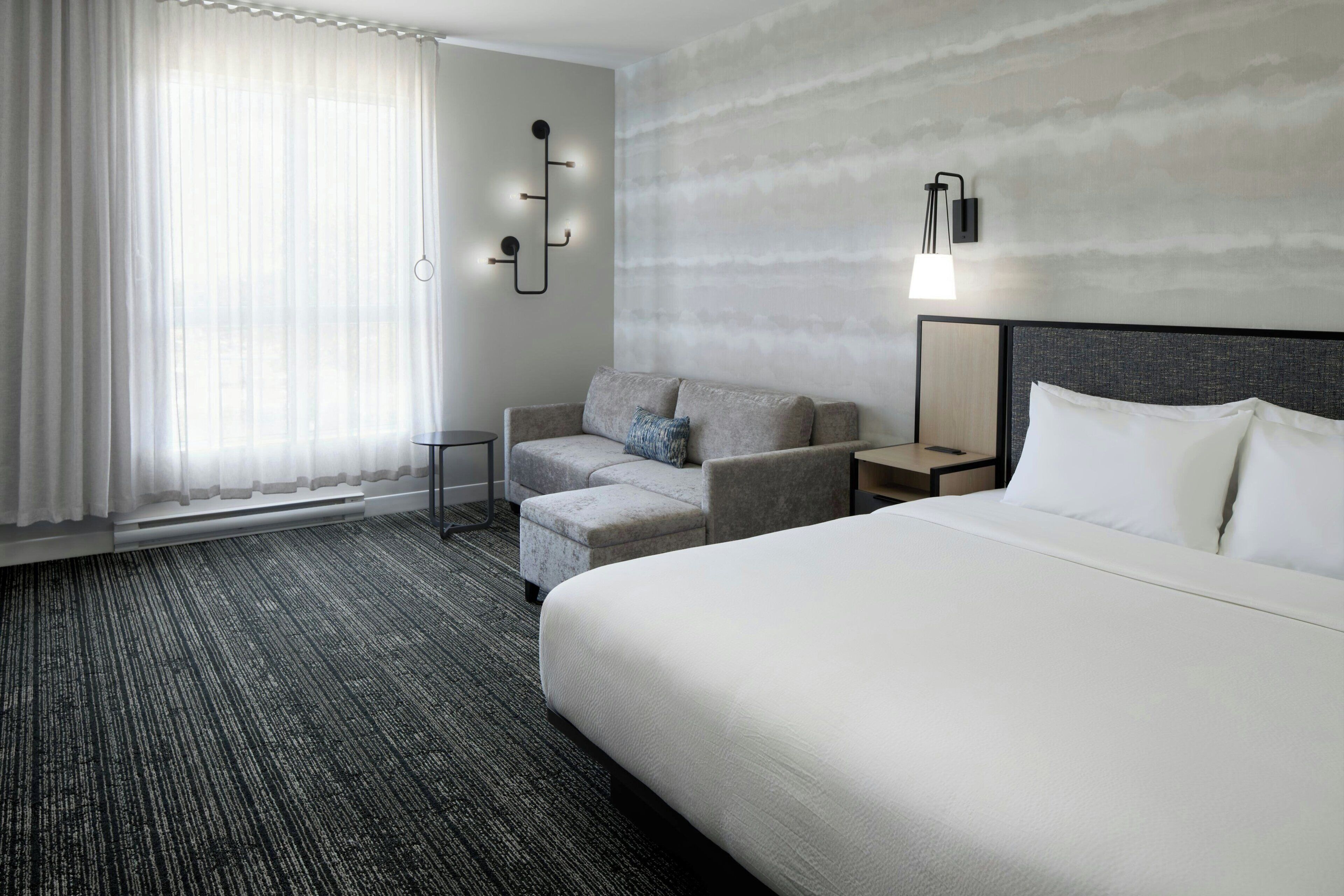 Quarto de hotel com uma cama grande, um sofá, uma mesinha e um candeeiro fixado na parede.