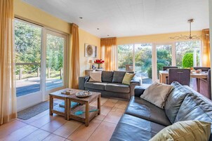 Uma sala de estar ampla com sofá, mesa de centro e área de jantar com mesa e cadeiras. Grandes janelas oferecem vista para o exterior.
