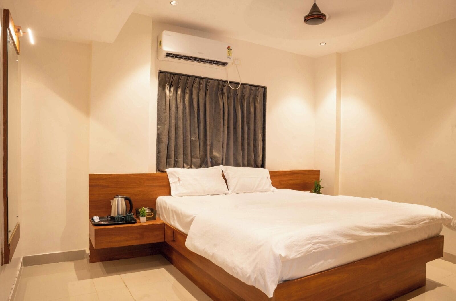 Quarto de hotel moderno com cama de madeira, mesinhas de cabeceira com chaleira e uma janela grande com cortinas.