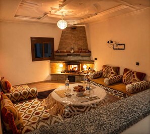 Uma sala de estar acolhedora com lareira, sofás estampados, uma mesa de centro redonda com petiscos e uma parede de pedra.
