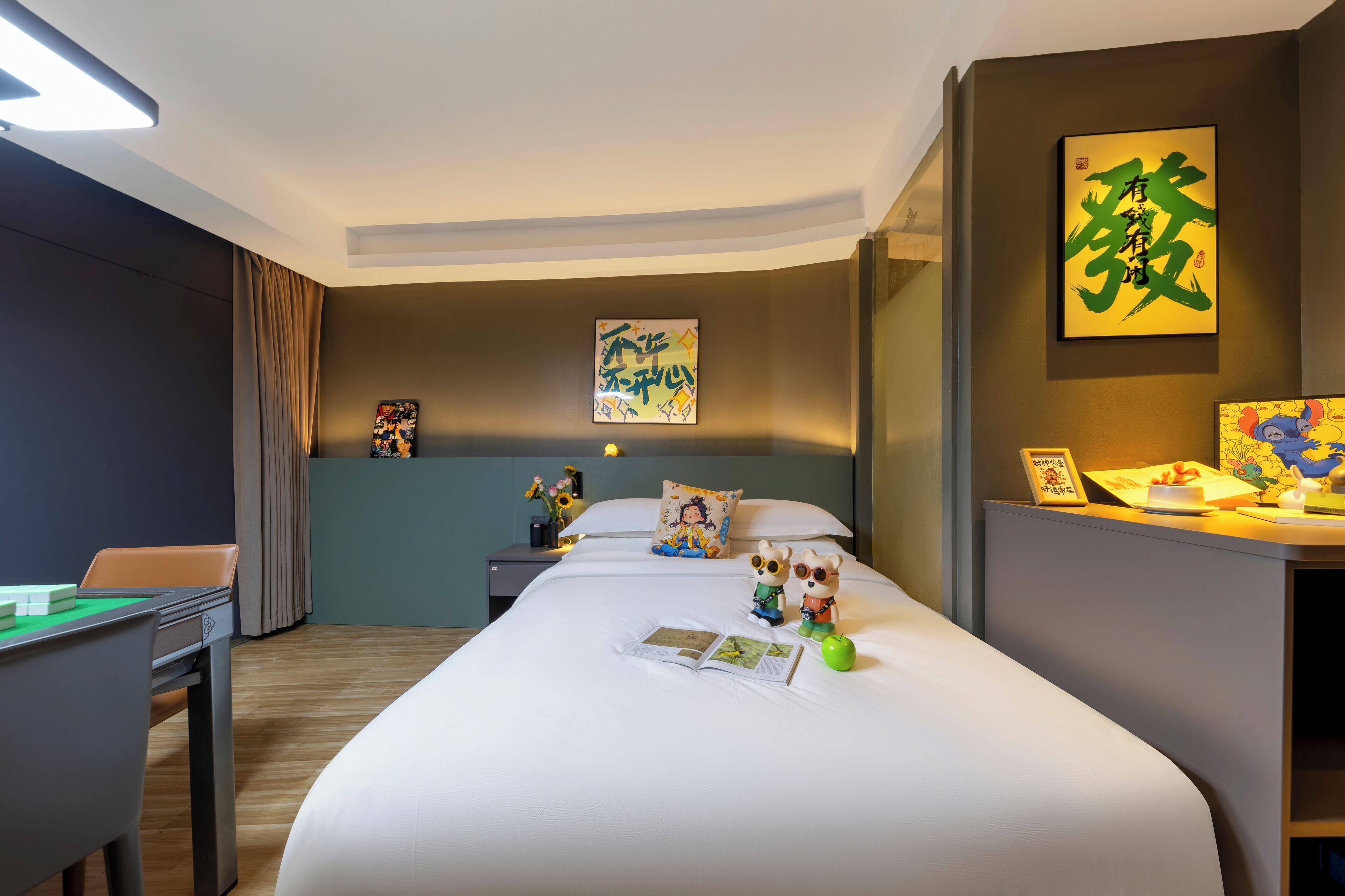 Quarto de hotel com cama bem arrumada, escrivaninha com jogo de tabuleiro verde, mesa de cabeceira com abajur e obras de arte nas paredes.