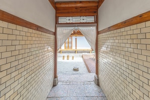 Um corredor com paredes revestidas de azulejos e teto de madeira, que leva a um edifício tradicional com porta de madeira e vista para um jardim.