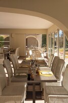 Uma área de refeições com uma mesa comprida posta para seis pessoas, rodeada por cadeiras de vime, com vista para o exterior através de grandes janelas.