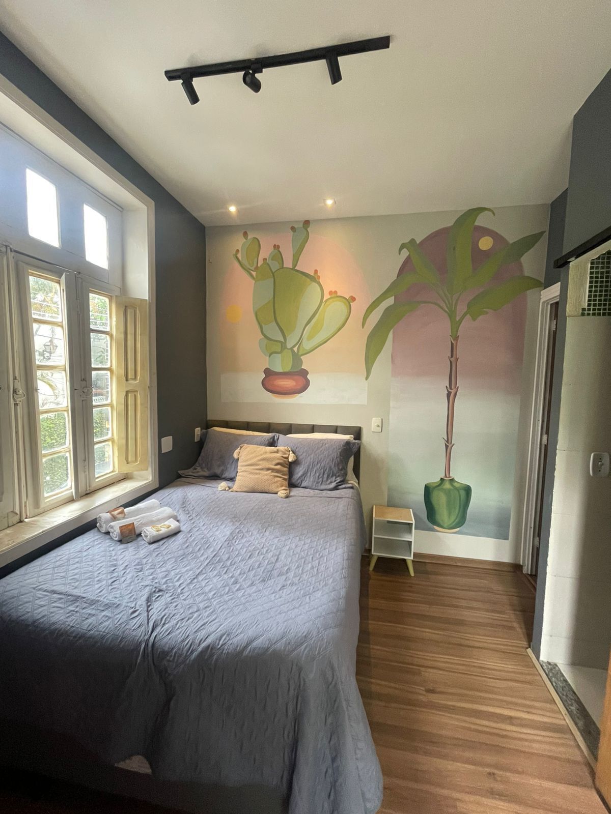 Quarto com cama, travesseiros, mesa de cabeceira, janela com cortinas e um mural na parede com cactos e plantas.
