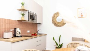 Cozinha moderna com micro-ondas, torradeira e cafeteira. Há uma área de refeições com uma mesa posta para dois e uma cadeira de vime.
