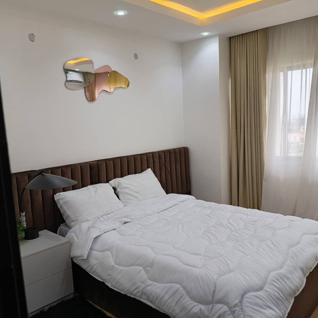 Quarto de hotel moderno com uma cama grande, uma mesinha de cabeceira, luminária fixada na parede e uma janela com cortinas.