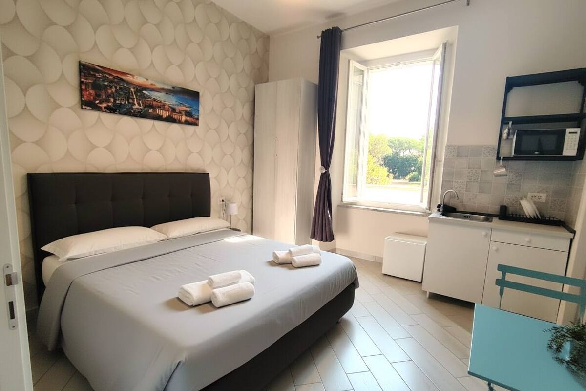 Quarto de hotel com cama, janela com cortinas, cozinha compacta e uma pequena mesa com cadeira.