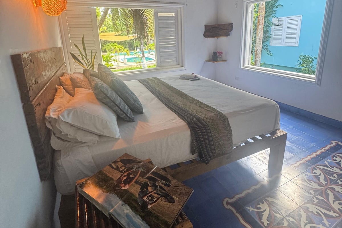 Quarto com cama, mesa de cabeceira, janela com vista para uma piscina e teto com vigas de madeira.
