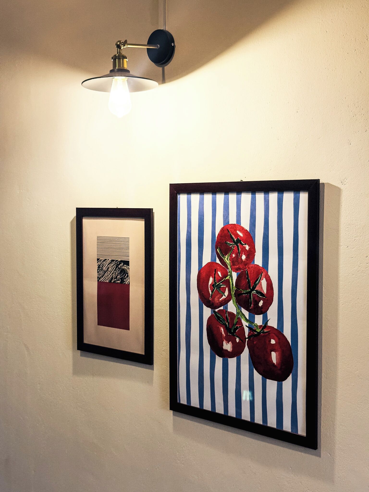 Duas obras de arte emolduradas numa parede, uma com um design abstrato em vermelho e preto, e outra com tomates vermelhos num fundo listrado em azul e branco.