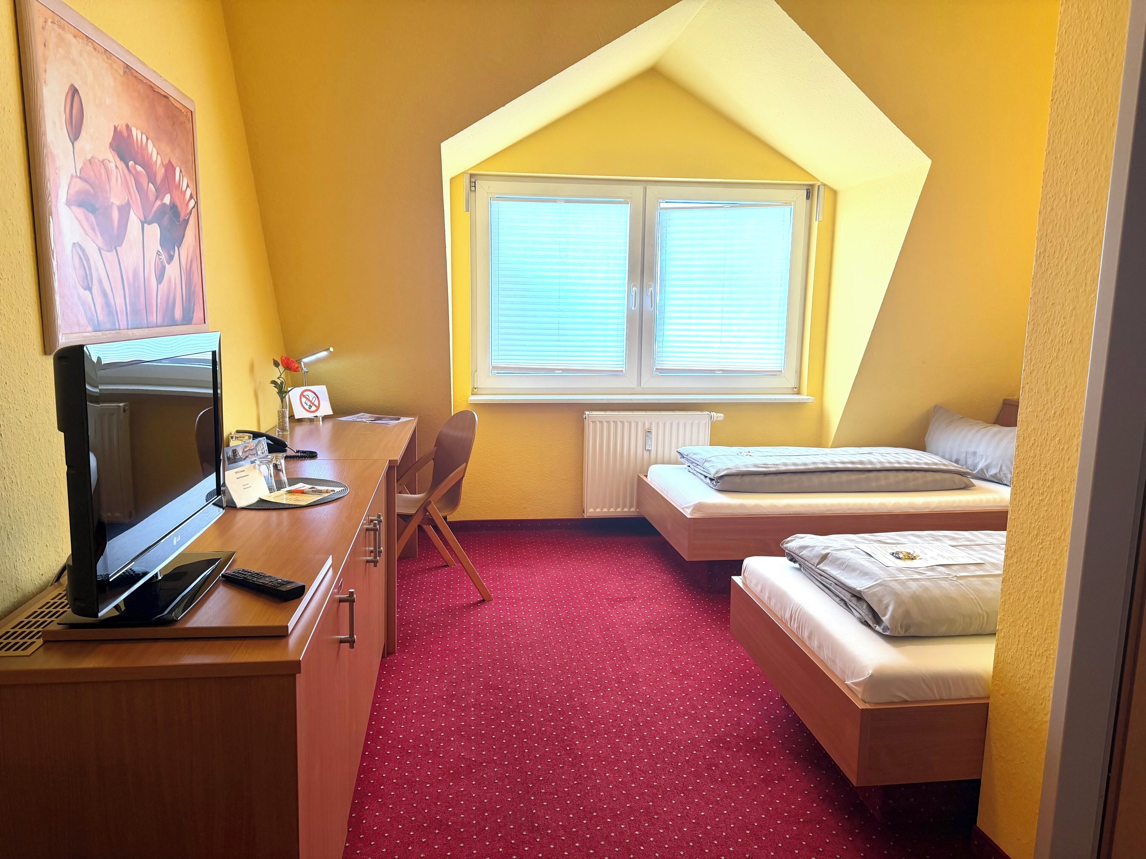 Um quarto pequeno e triangular com uma cama, uma escrivaninha com computador e uma janela com persianas.