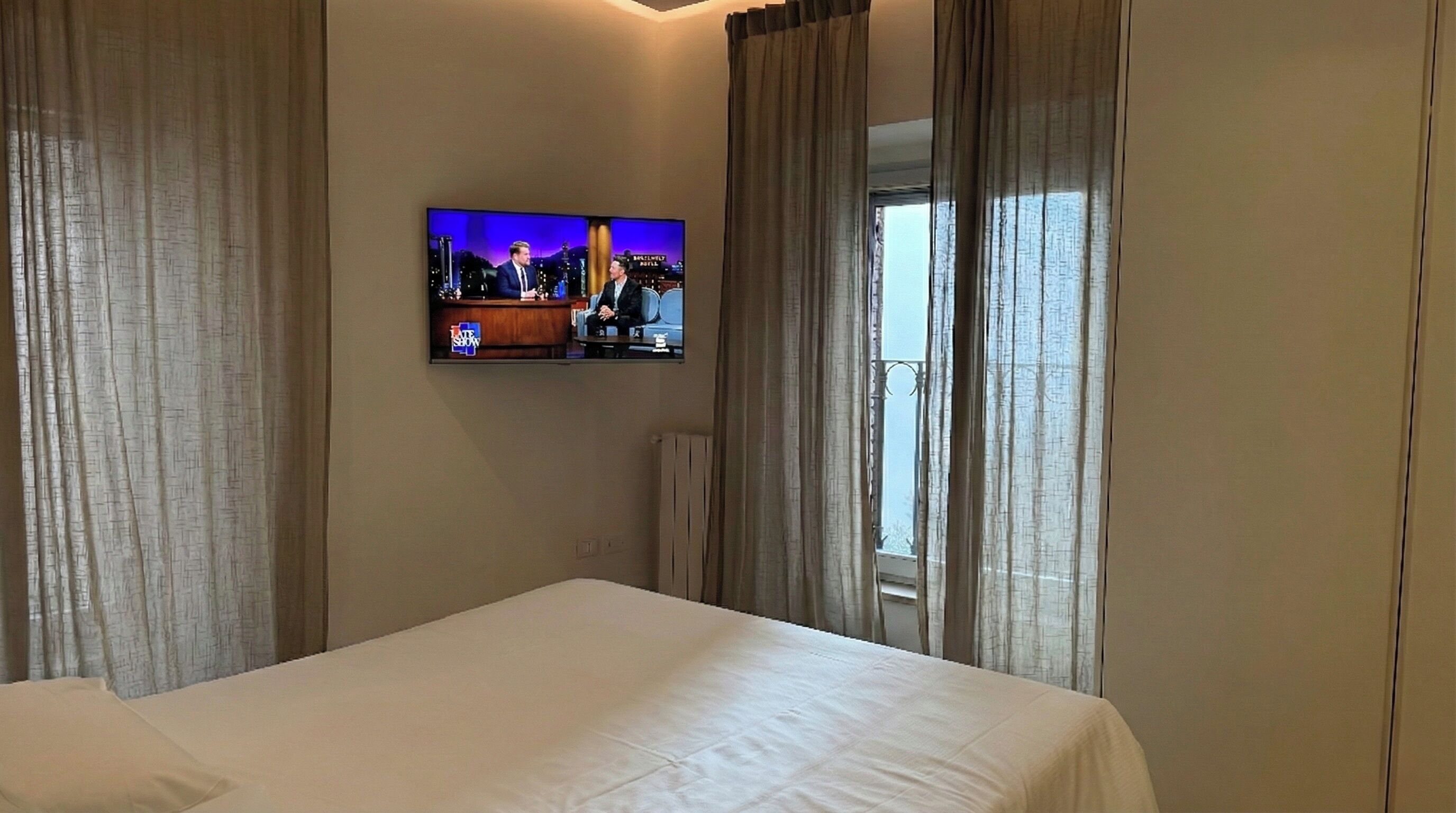 Quarto de hotel com uma cama, televisão fixada na parede e uma janela com cortinas.