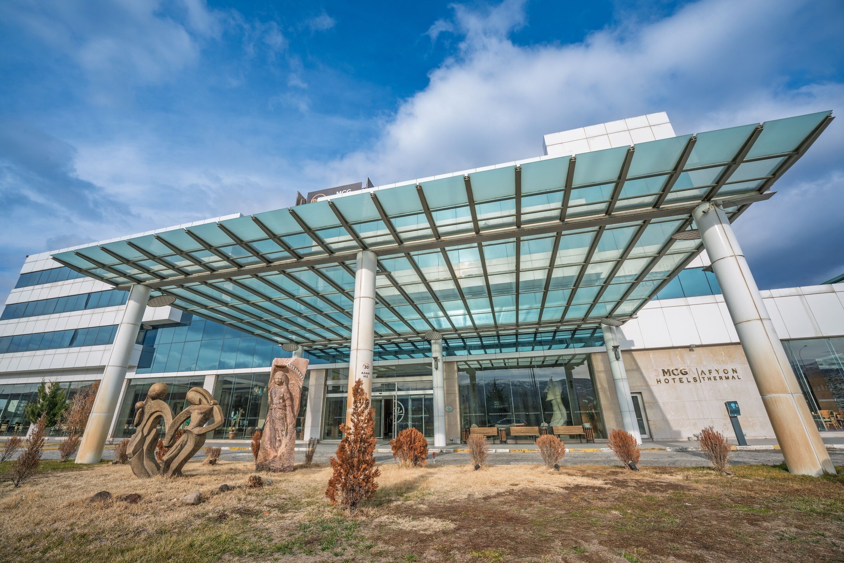 Hotel moderno com fachada de vidro, entrada coberta e uma escultura em primeiro plano.