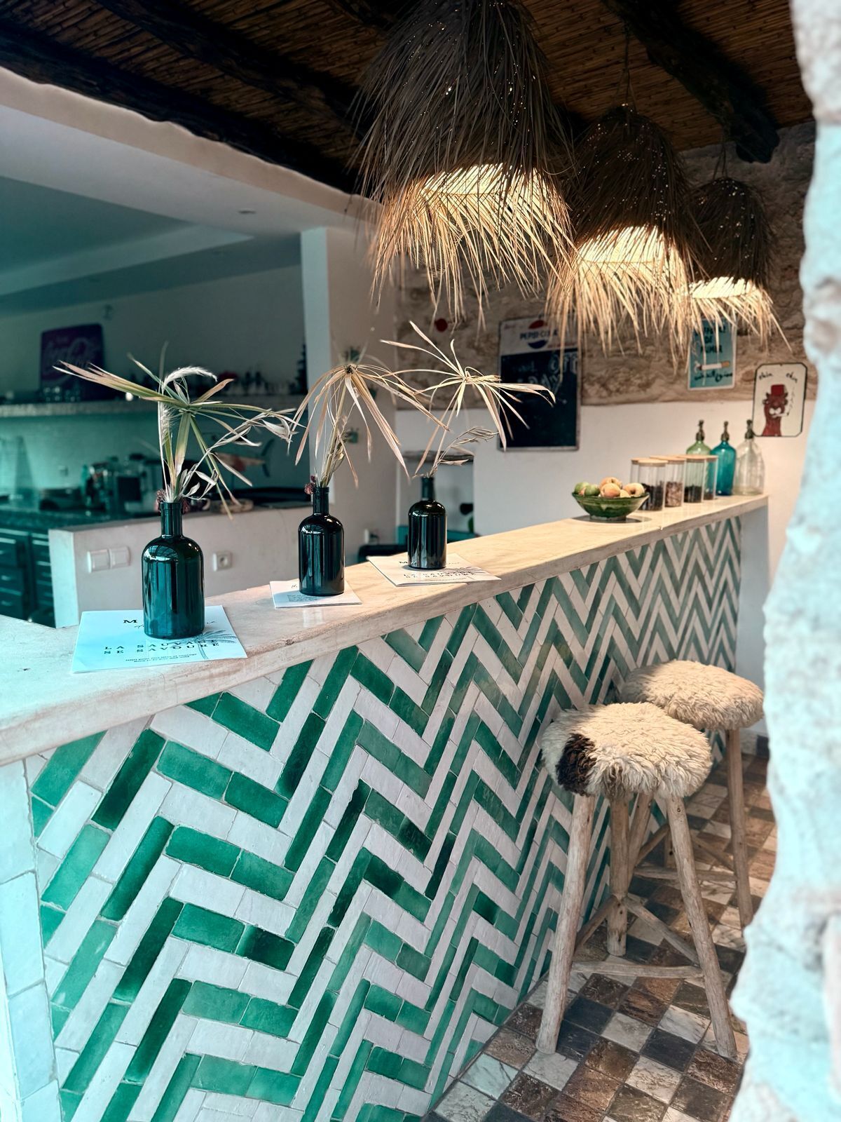 Uma área de bar com painel de azulejos em padrão Chevron, folhagens de palmeira penduradas e um banquinho de madeira.