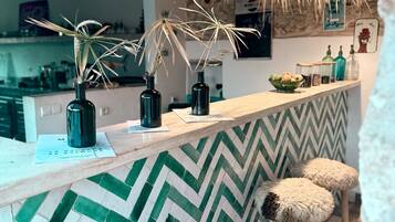 Uma área de bar com painel de azulejos em padrão Chevron, folhagens de palmeira penduradas e um banquinho de madeira.