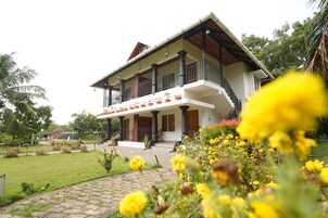 Um edifício de dois andares com varanda, cercado por um jardim com flores amarelas.