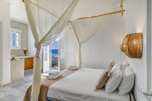 Um quarto com cama, cortinas brancas, um cesto de vime e vista para o mar.